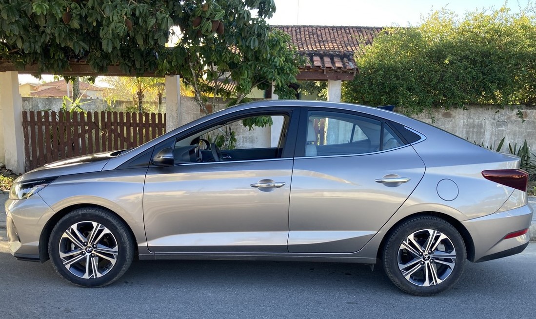 Foto mostrando a lateral esquerda de um Hyundai HB20S 2023 cinza estacionado em frente a uma casa.